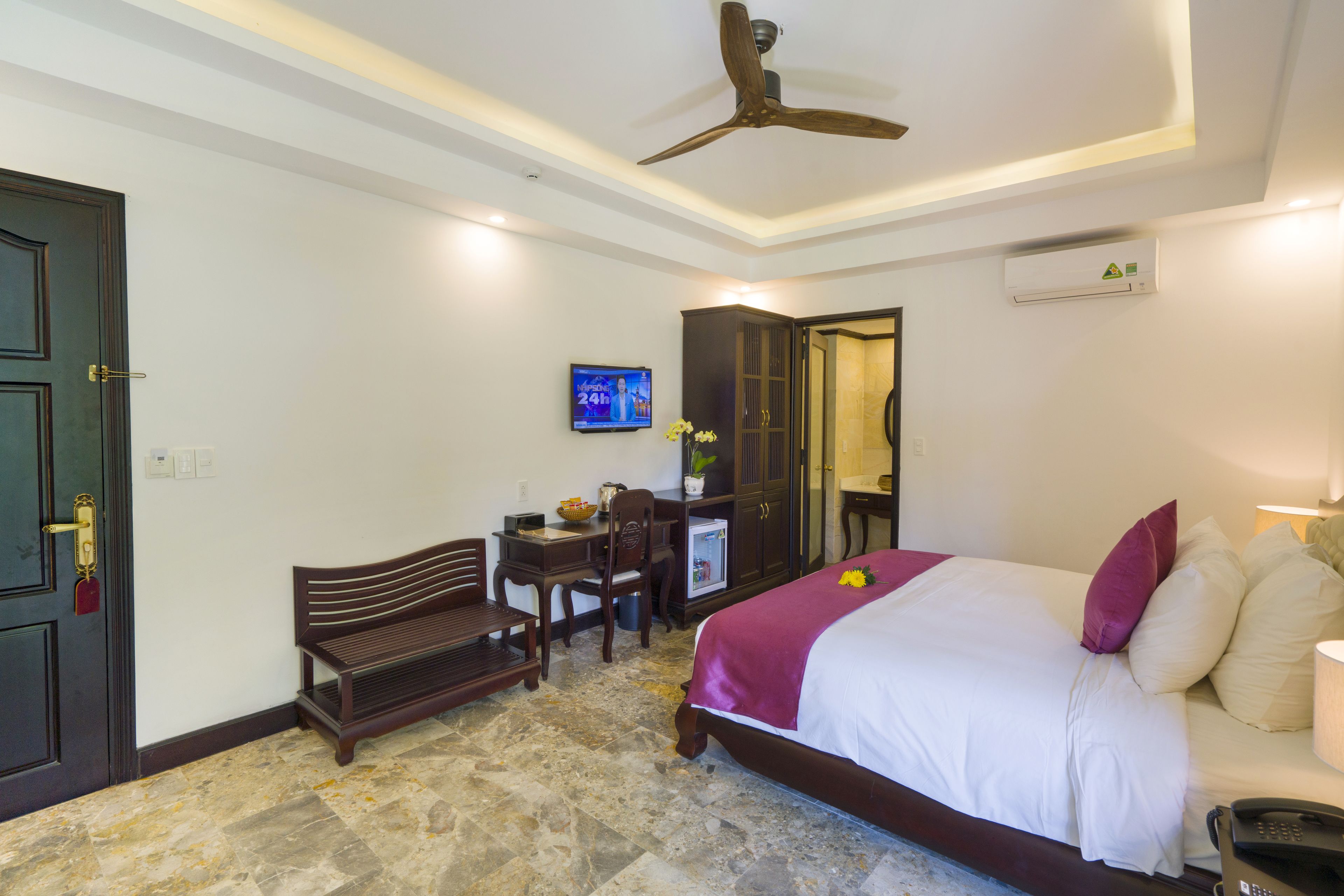 Quarto de hotel com cama, escrivaninha, cadeira, televisão e ventilador de teto.