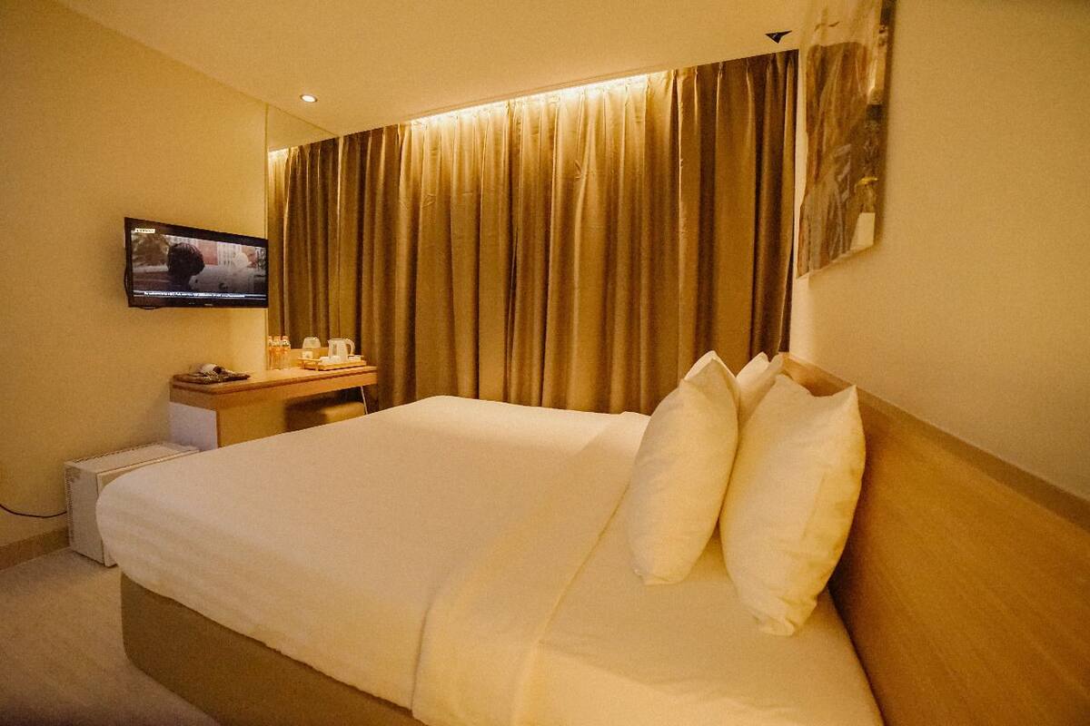 Quarto de hotel com uma cama grande, televisão de tela plana na parede, escrivaninha com telefone e cafeteira, e janela com cortinas.