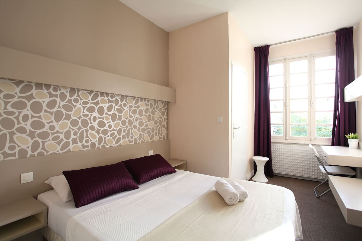 Quarto de hotel com cama, escrivaninha e janela com cortinas.