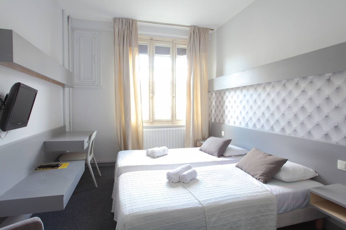 Quarto de hotel com duas camas, uma mesa, uma televisão e uma janela com cortinas.