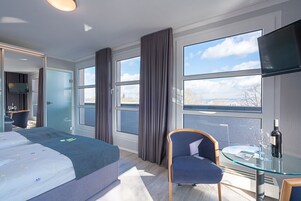 Quarto de hotel com uma cama, uma cadeira, uma mesa de vidro com uma garrafa e copos, e uma televisão na parede.