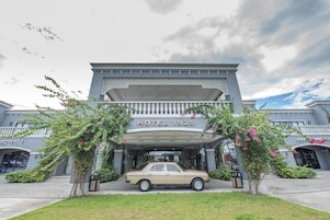 Entrada de hotel com um carro antigo estacionado na frente, rodeada por vegetação e um céu limpo.
