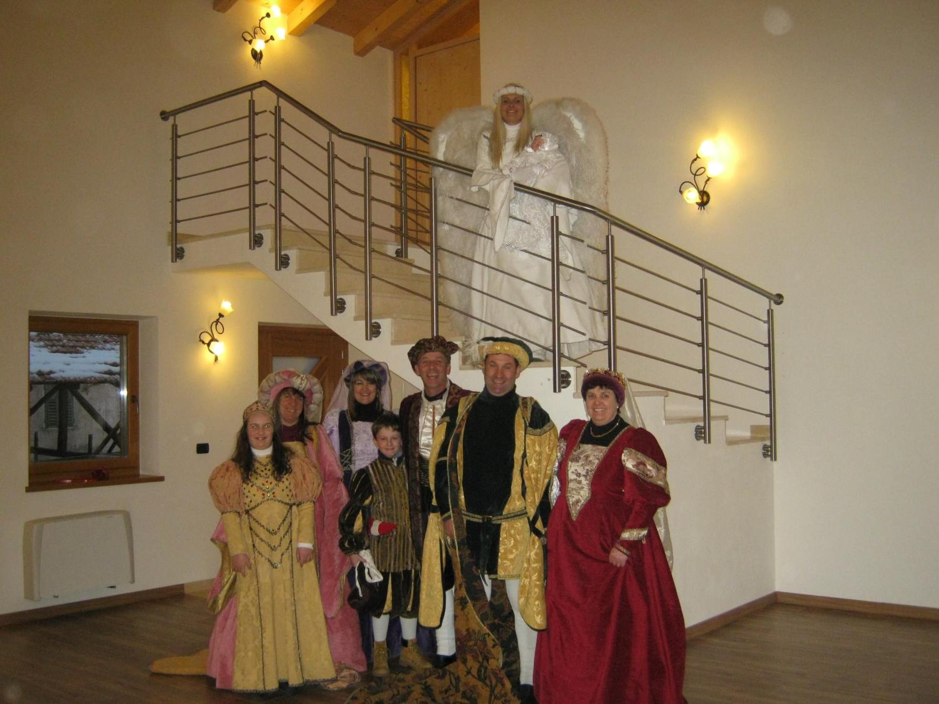 Um grupo de pessoas com trajes históricos posando em um quarto com escada e janela com vista para uma paisagem coberta de neve.