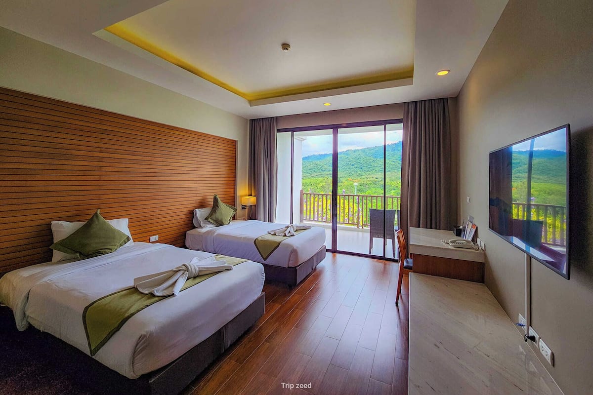 Quarto de hotel com duas camas, varanda com vista panorâmica, televisão de tela plana e cabeceira de madeira.