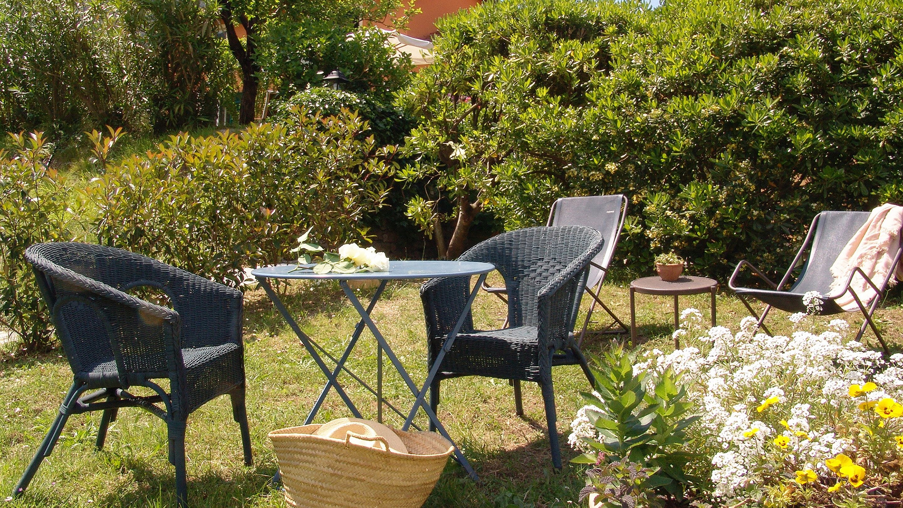 Um jardim com móveis de exterior, incluindo duas cadeiras e uma mesa, rodeado por vegetação exuberante e flores.
