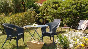 Um jardim com móveis de exterior, incluindo duas cadeiras e uma mesa, rodeado por vegetação exuberante e flores.
