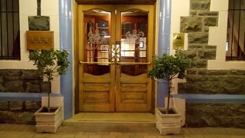 Entrada de hotel com portas de madeira dupla, uma placa e dois vasos de plantas.
