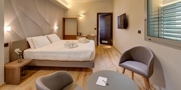 Quarto de hotel moderno com uma cama bem arrumada, um espaço de estar com uma poltrona e uma pequena mesa, e uma grande janela com persianas.