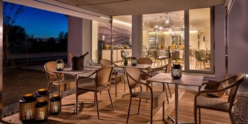 Uma área de refeições ao ar livre com cadeiras e mesas de vime, um bar com balcão e banquetas, e vista para o espaço de refeições interno.