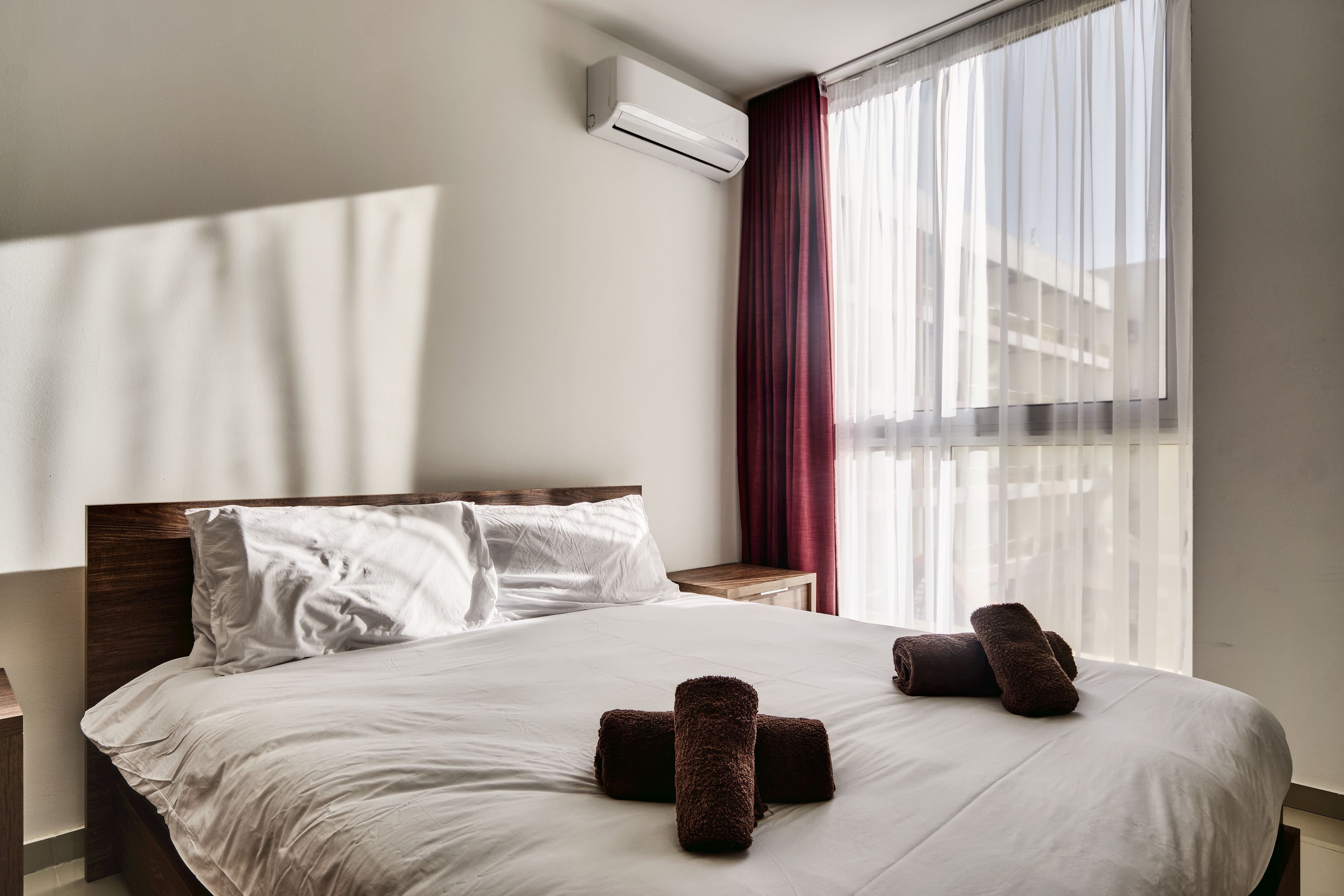 Quarto de hotel com uma cama grande, roupa de cama branca, cabeceira de madeira, mesa de cabeceira, janela com cortinas leves e ar condicionado.