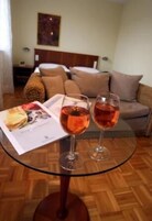 Quarto de hotel com uma cama, um sofá e uma mesa de vidro com duas taças de vinho e uma revista.