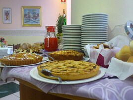 Uma mesa de buffet com diversos produtos de panificação, incluindo tortas e doces, e uma pilha de pratos.