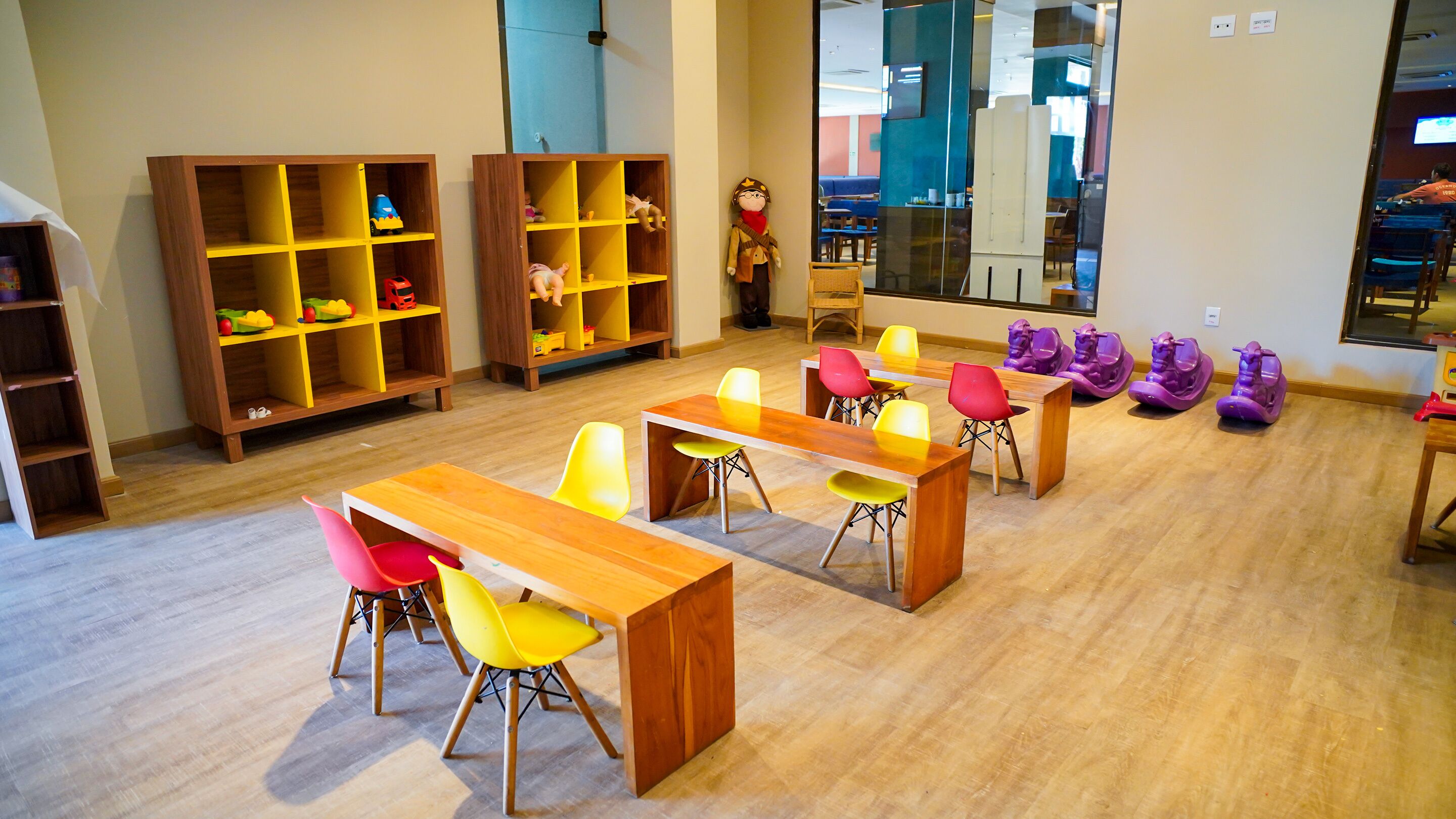 Espaço infantil com mesas de madeira e cadeiras coloridas, cercado por prateleiras com brinquedos.
