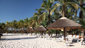Uma praia com guarda-sóis de palha, espreguiçadeiras brancas e palmeiras.