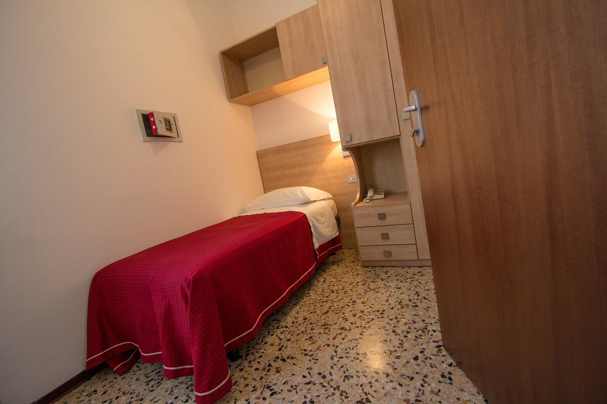 Um quarto pequeno e aconchegante com uma cama de solteiro, uma mesinha de cabeceira, um guarda-roupa de madeira e uma prateleira fixada na parede.