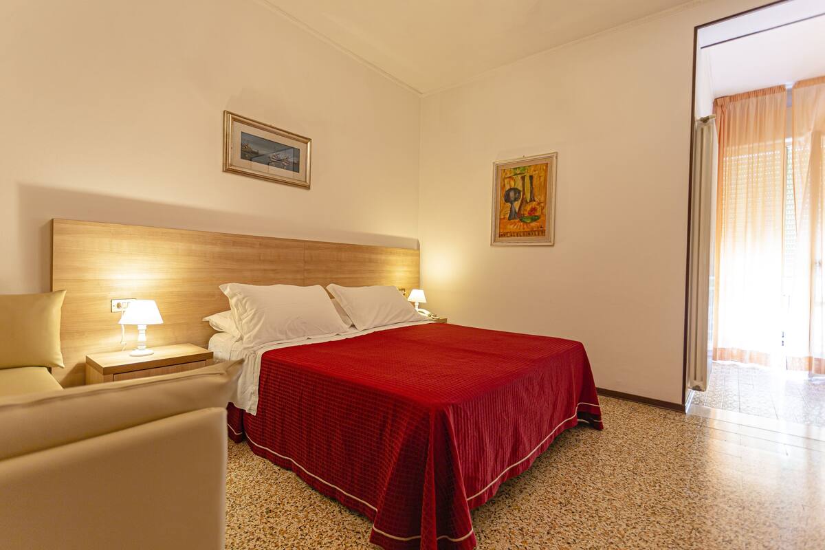 Quarto de hotel com cabeceira de madeira, colcha vermelha, poltrona bege e duas luminárias de mesa.
