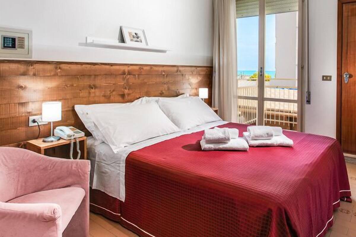 Quarto de hotel com cabeceira de madeira, cama com colcha vermelha, mesa de cabeceira com luminária, telefone e uma cadeira.
