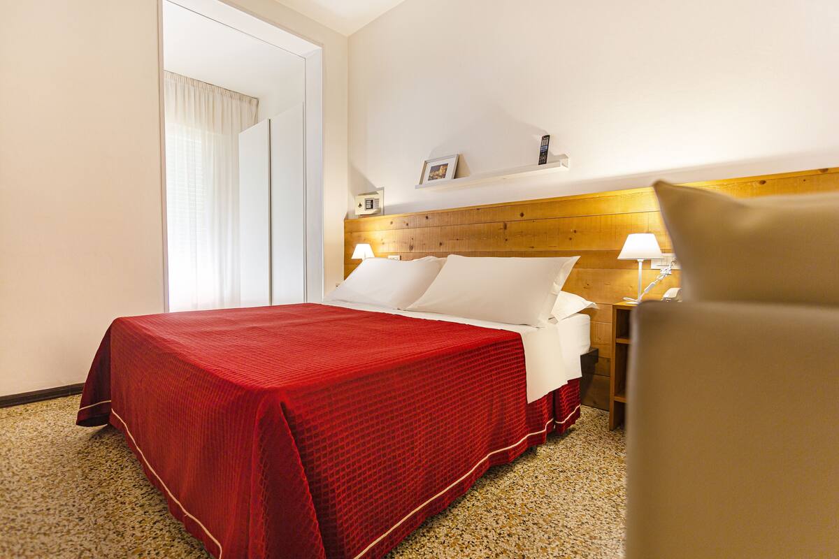 Quarto de hotel com uma cama grande, cabeceira de madeira, mesa de cabeceira com abajur e uma cadeira.