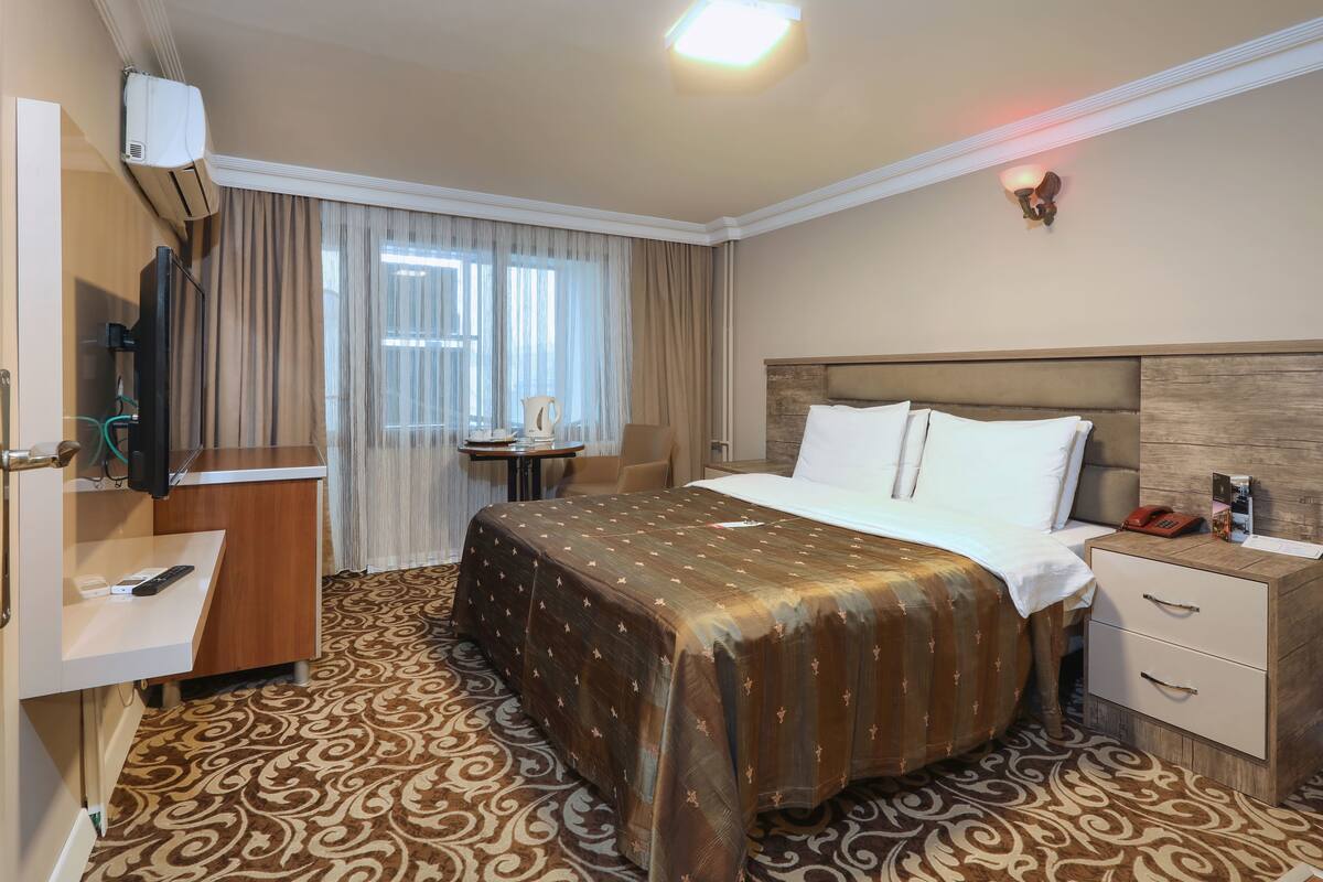 Quarto de hotel com uma cama grande, uma mesa de cabeceira com abajur, uma televisão e uma escrivaninha com cadeira.