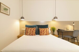 Quarto de hotel com uma cama grande, duas almofadas estampadas, uma escrivaninha com cadeira e uma pequena mesa com uma planta.