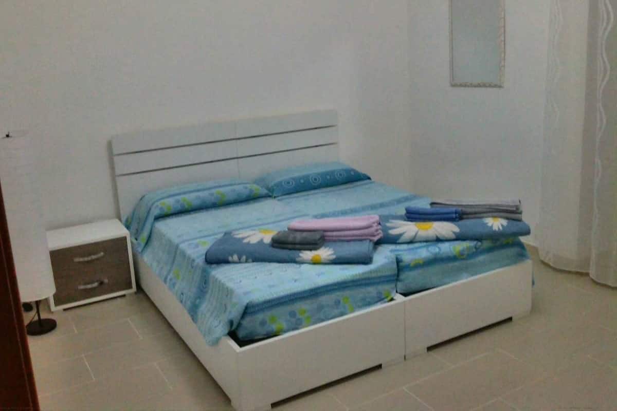Uma cama bem arrumada com colcha floral, uma mesinha de cabeceira com abajur e um pequeno móvel com gavetas.