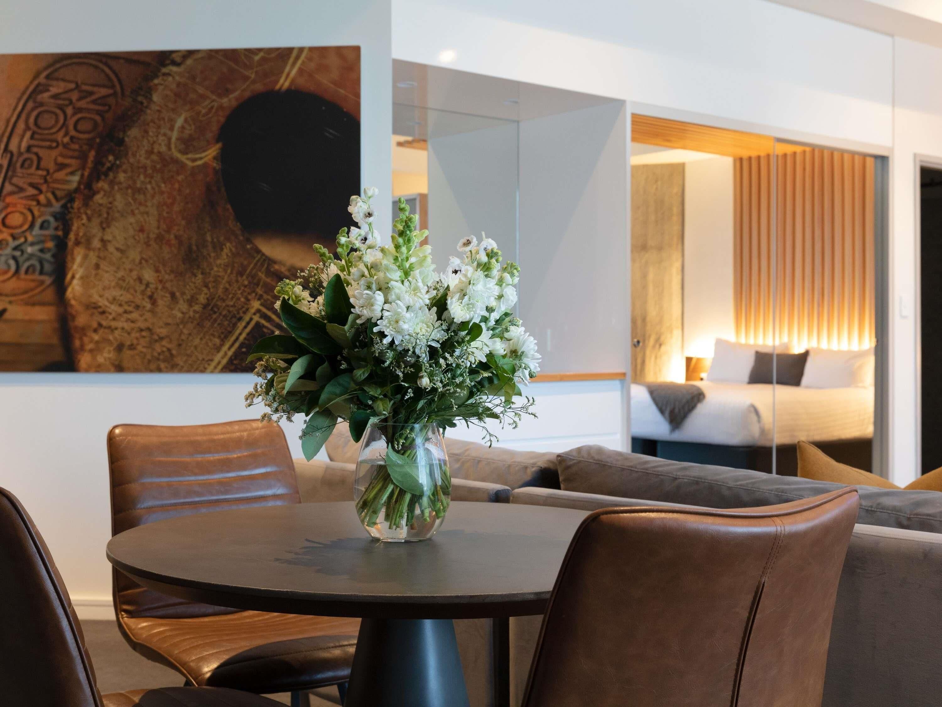 Quarto de hotel moderno com área de jantar, com uma mesa redonda e cadeiras de couro, um vaso decorativo com flores brancas e um grande quadro abstrato na parede.