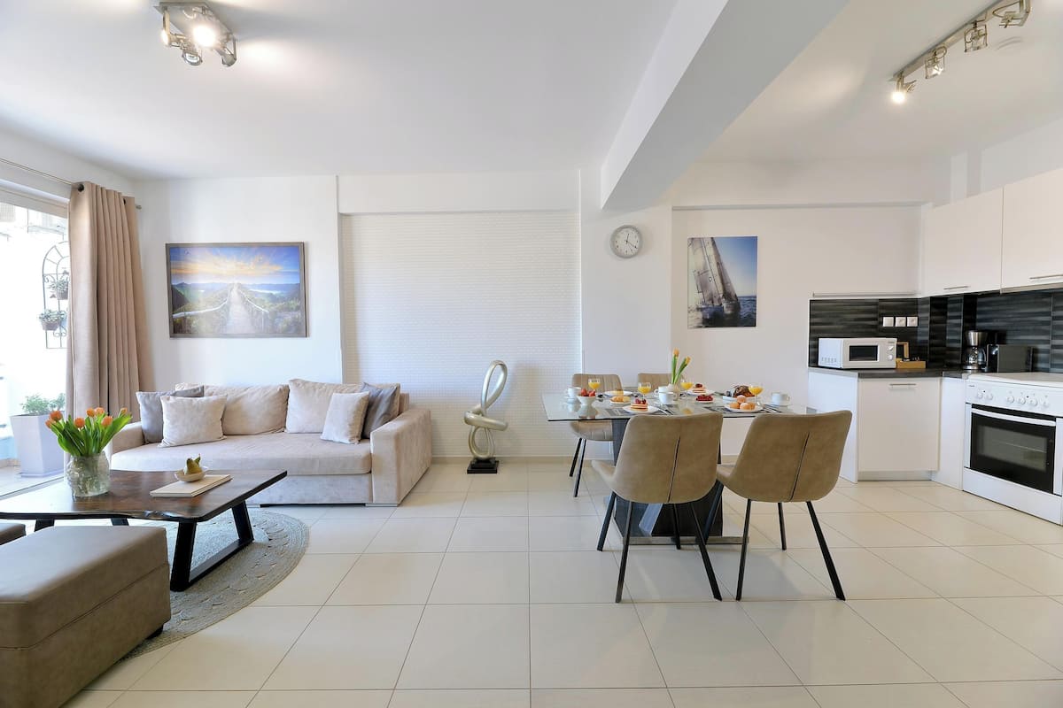 Sala moderna com sofá, mesa de centro e área de jantar com cadeiras. Cozinha com eletrodomésticos visível ao fundo.