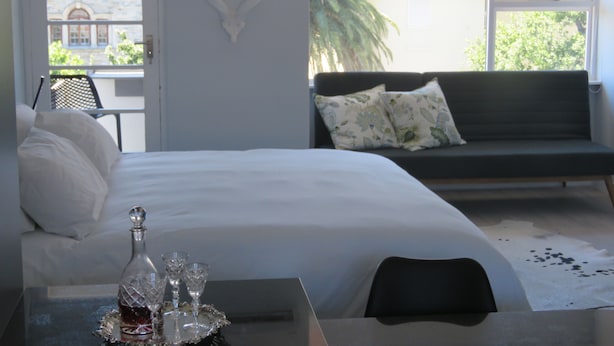 Quarto com cama, mesa com garrafa e copos, sofá com almofadas e vista para área verde lá fora.