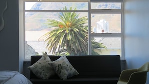 Quarto com uma janela ampla, um sofá com almofadas florais e vista para uma montanha e edifícios.