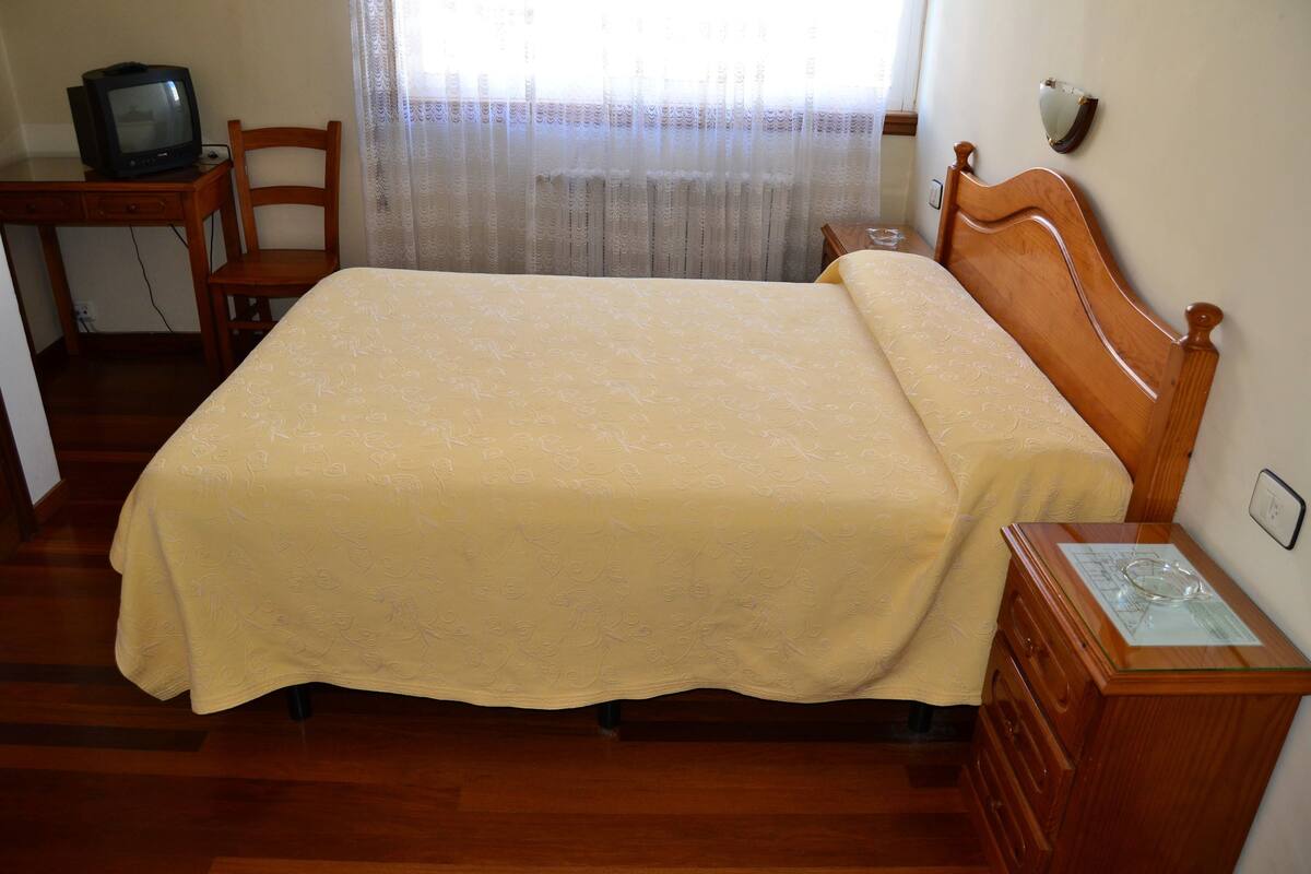 Quarto com cama, cabeceira de madeira, mesa de cabeceira, cômoda, televisão sobre uma escrivaninha e janela com cortinas.