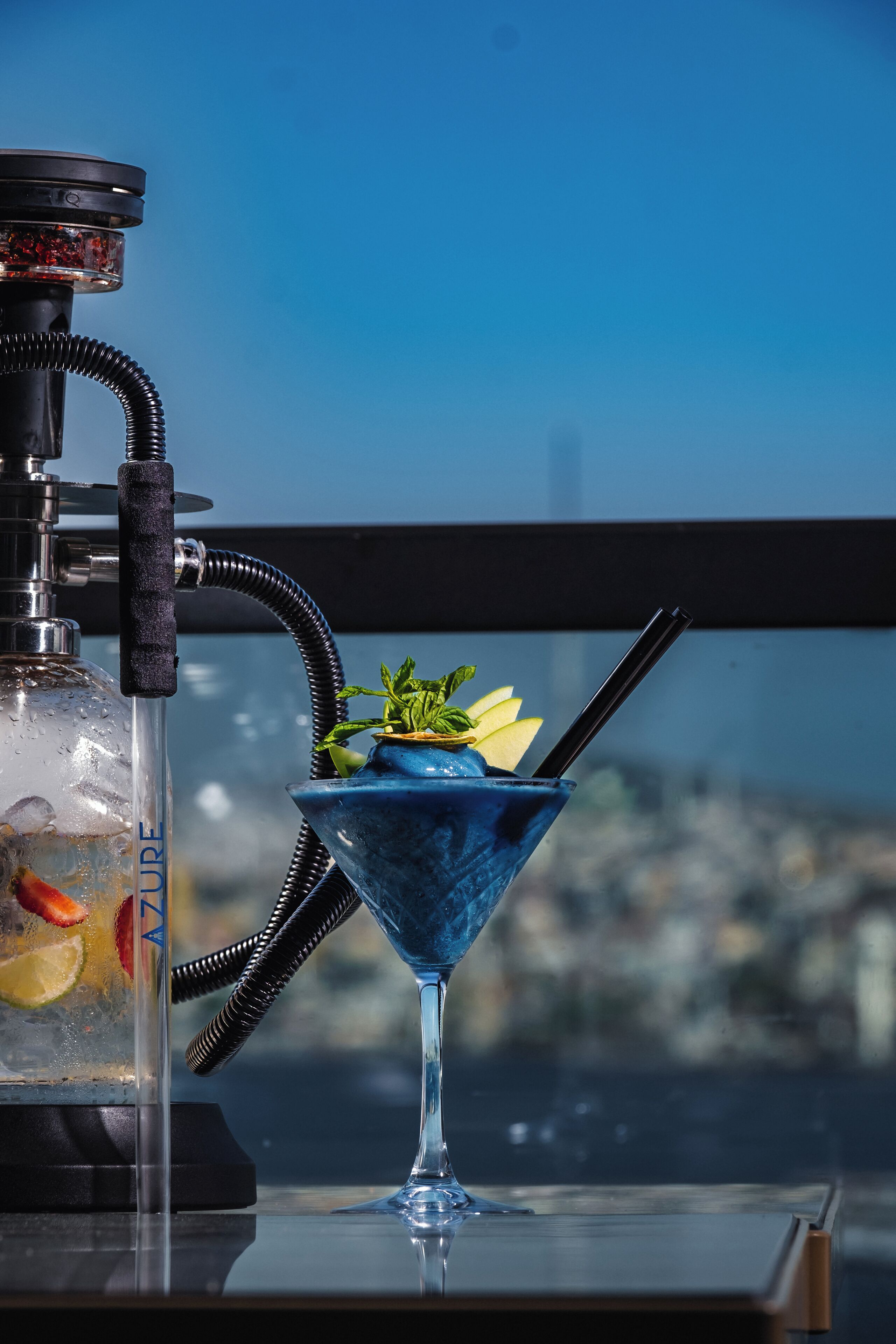 Um coquetel em copo de martini com canudo preto, decorado com folhas de hortelã e fatias de limão, ao lado de um narguilé com mangueira.
