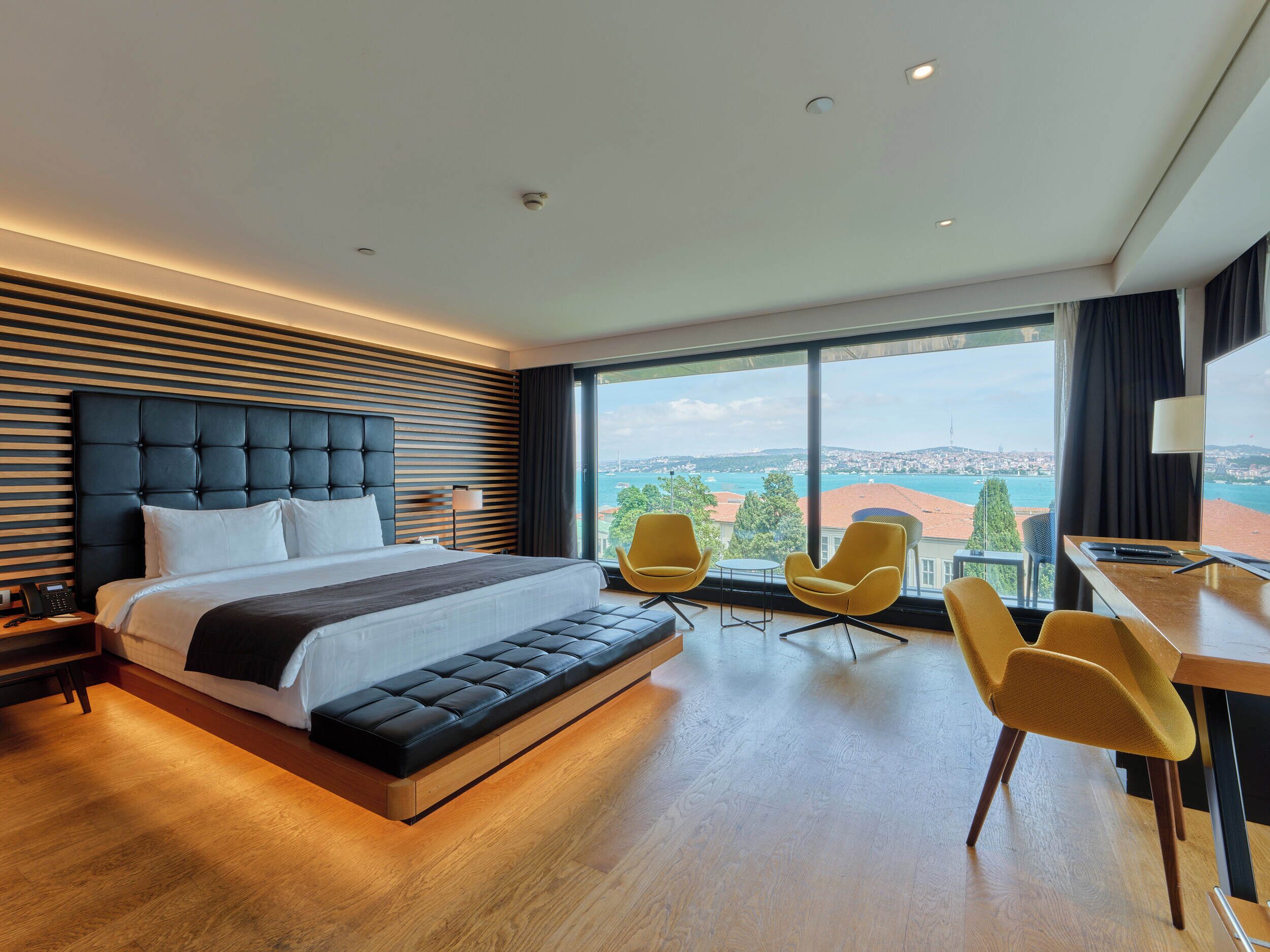 Quarto de hotel moderno com uma cama grande, uma escrivaninha com cadeiras e vista para a cidade.