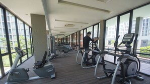 Academia com aparelhos de cardio, um homem usando uma elíptica, e grandes janelas com vista para edifícios e áreas verdes.