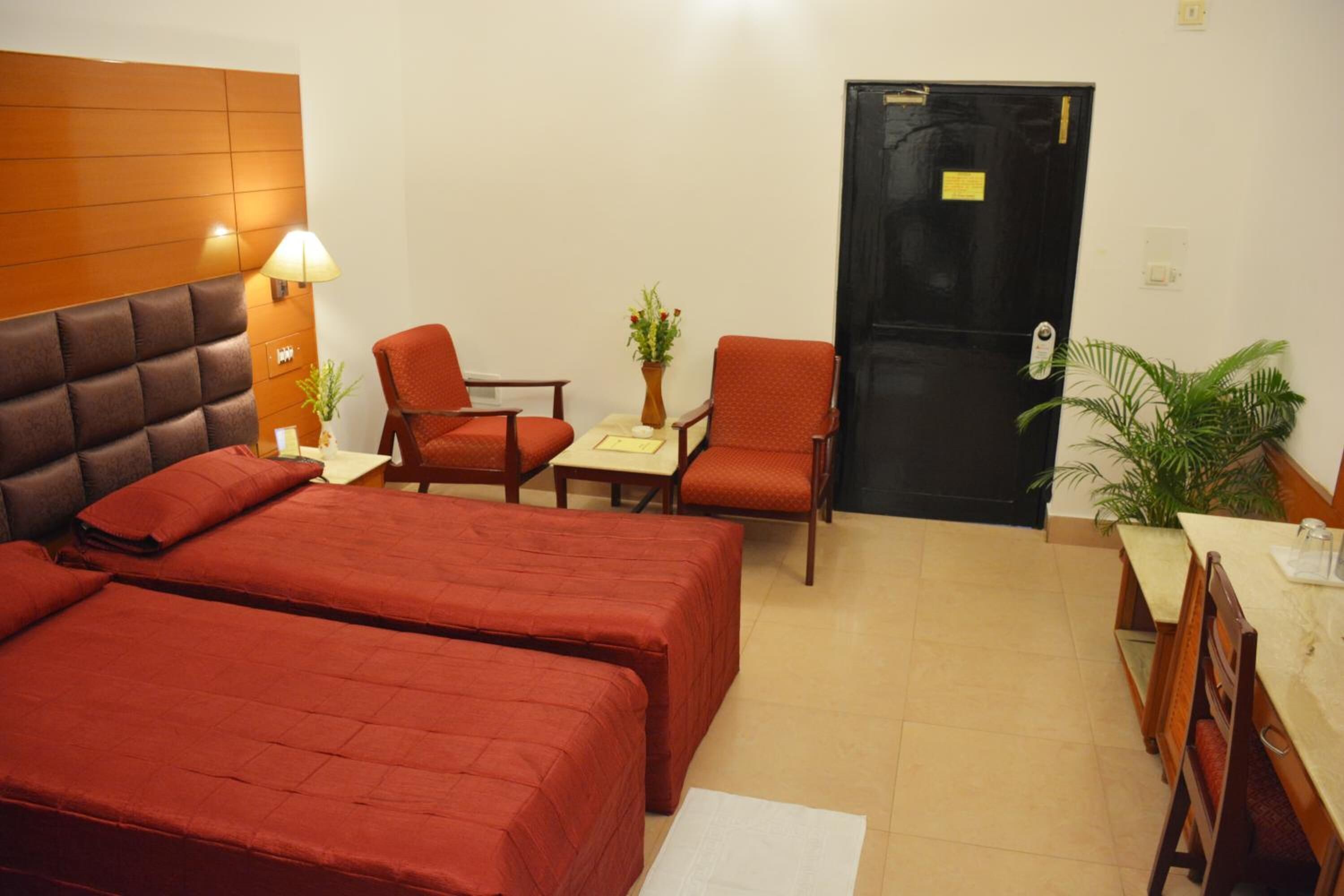 Quarto de hotel com duas camas, uma escrivaninha, duas cadeiras, uma porta e um vaso de planta.