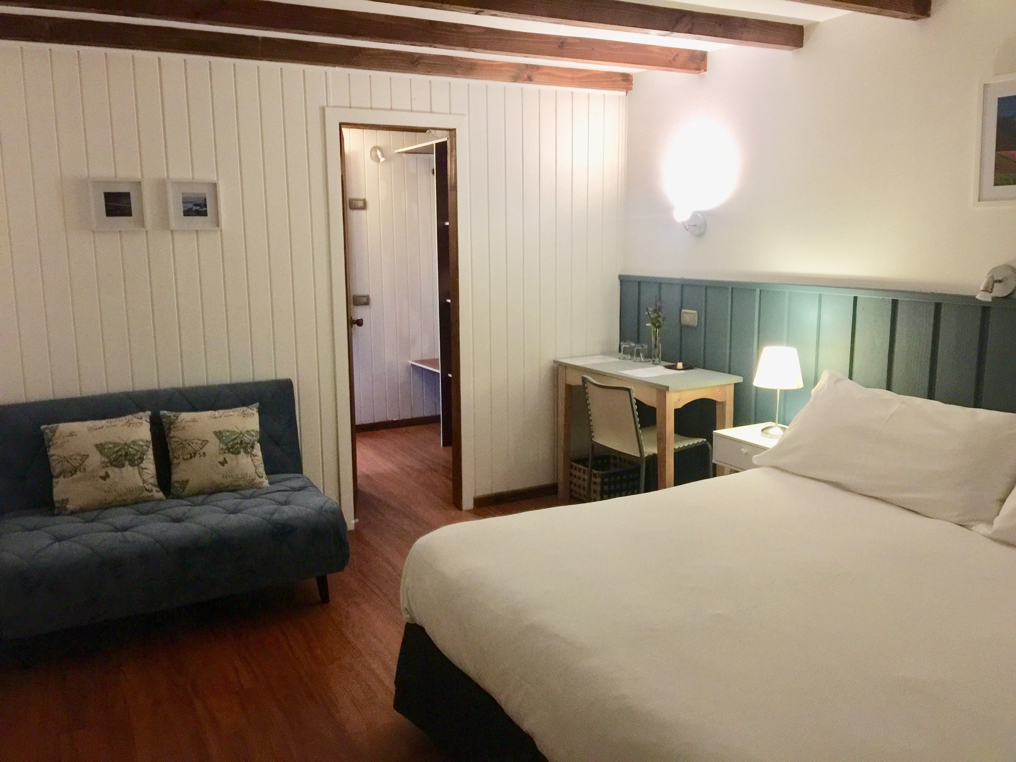 Quarto de hotel com uma cama, um sofá azul, uma escrivaninha com cadeira e uma mesa de cabeceira com abajur.