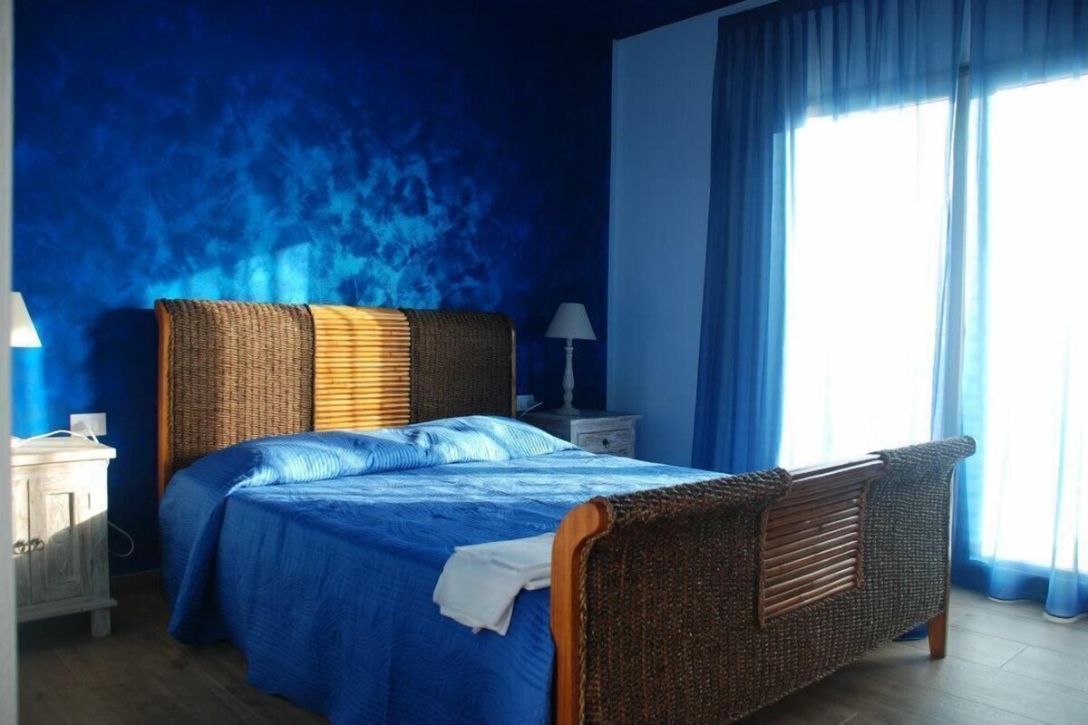 Quarto com cama, mesa de cabeceira, luminária e janela com cortinas.