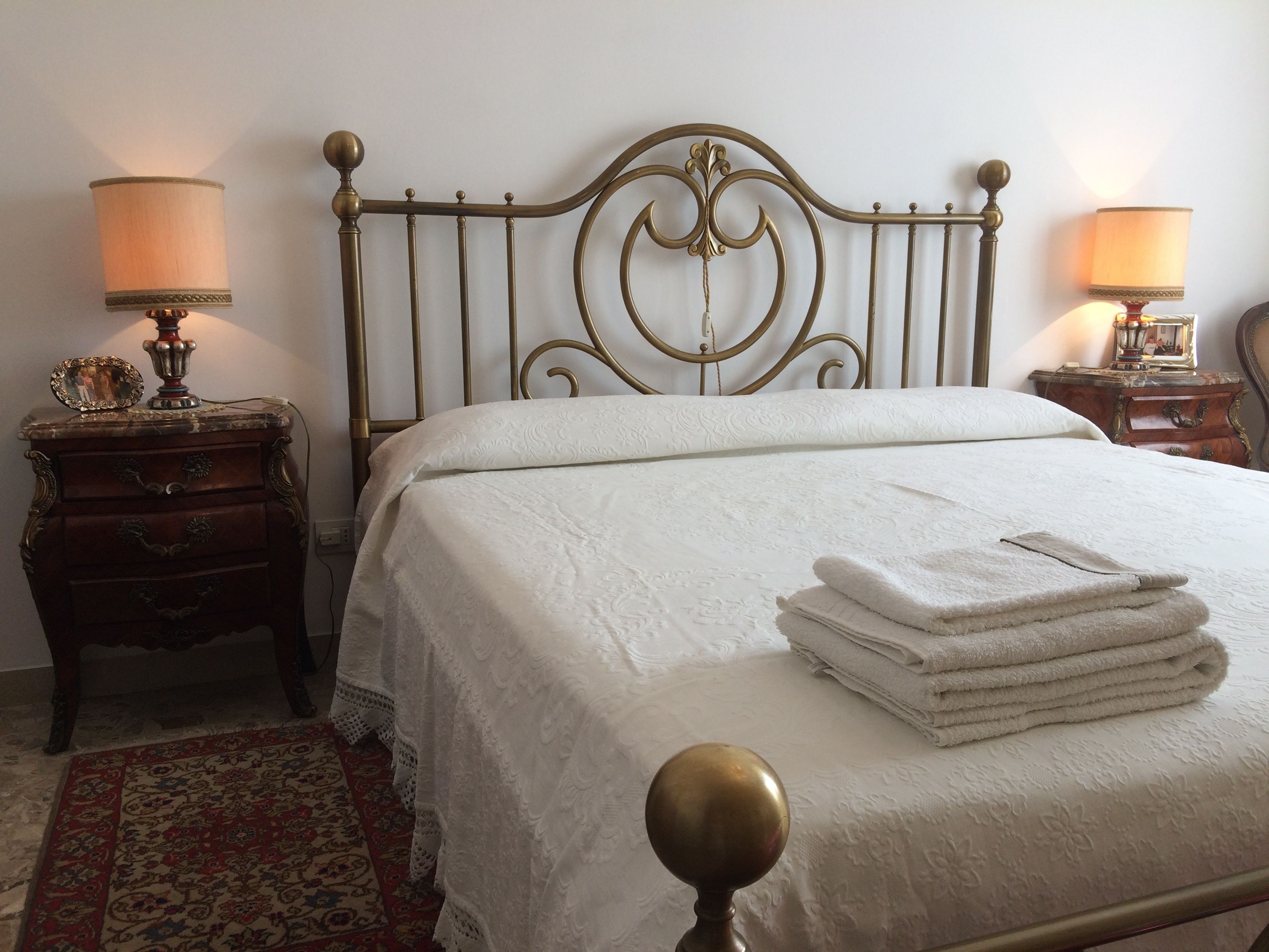 Cama bem arrumada com colcha branca e toalhas dobradas, com duas mesas de cabeceira com luminárias e um objeto decorativo em uma delas.