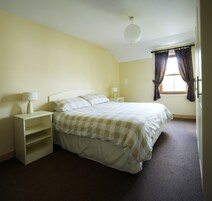 Quarto individual com uma colcha xadrez, mesa de cabeceira com luminária e janela com cortinas.