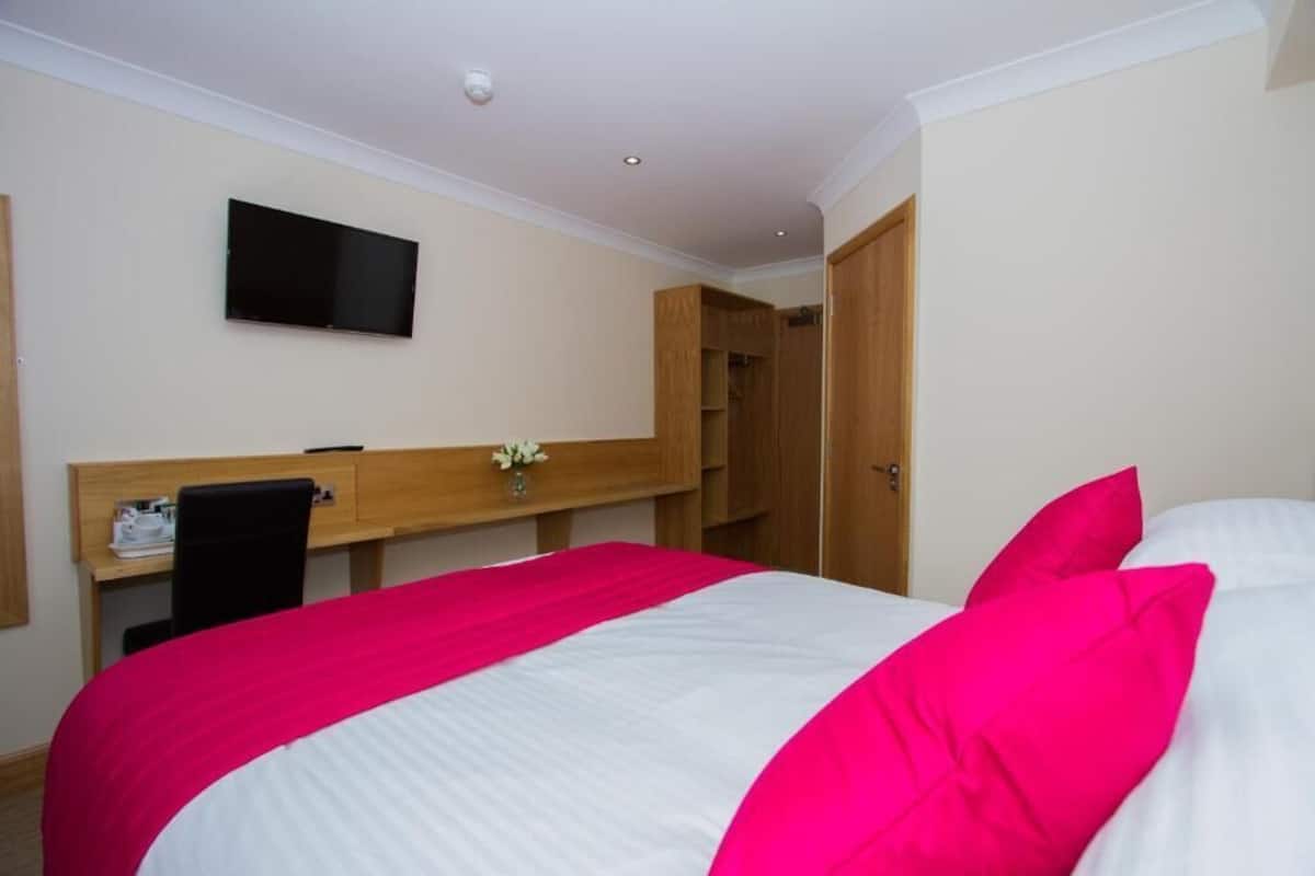 Quarto de hotel com cama, escrivaninha, cadeira, televisão e guarda-roupa.