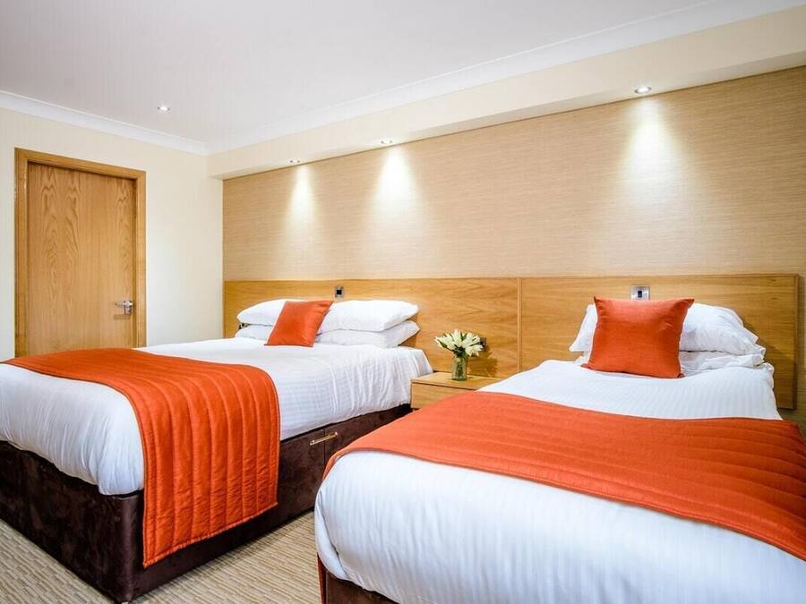 Quarto de hotel com duas camas, cada uma com cabeceira de madeira, roupa de cama branca e mantas laranja.
