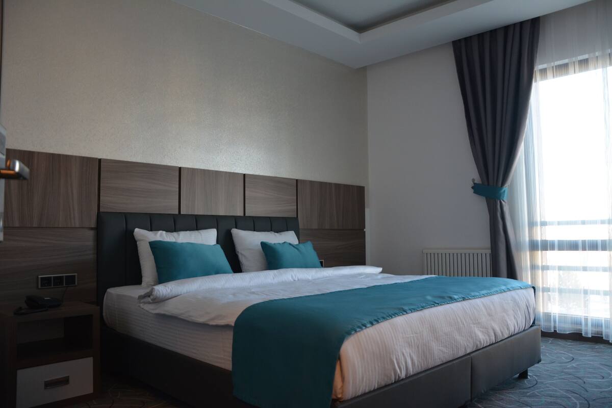 Quarto de hotel com cama, mesa de cabeceira, janela com cortinas e parede com textura.