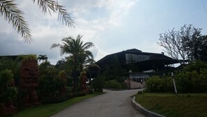 Uma trilha leva a um edifício moderno com fachada de vidro, cercado por plantas tropicais e estátuas.