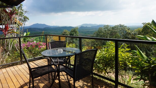 Um deck de madeira com mesa e cadeiras de vidro, com vista para uma paisagem montanhosa.