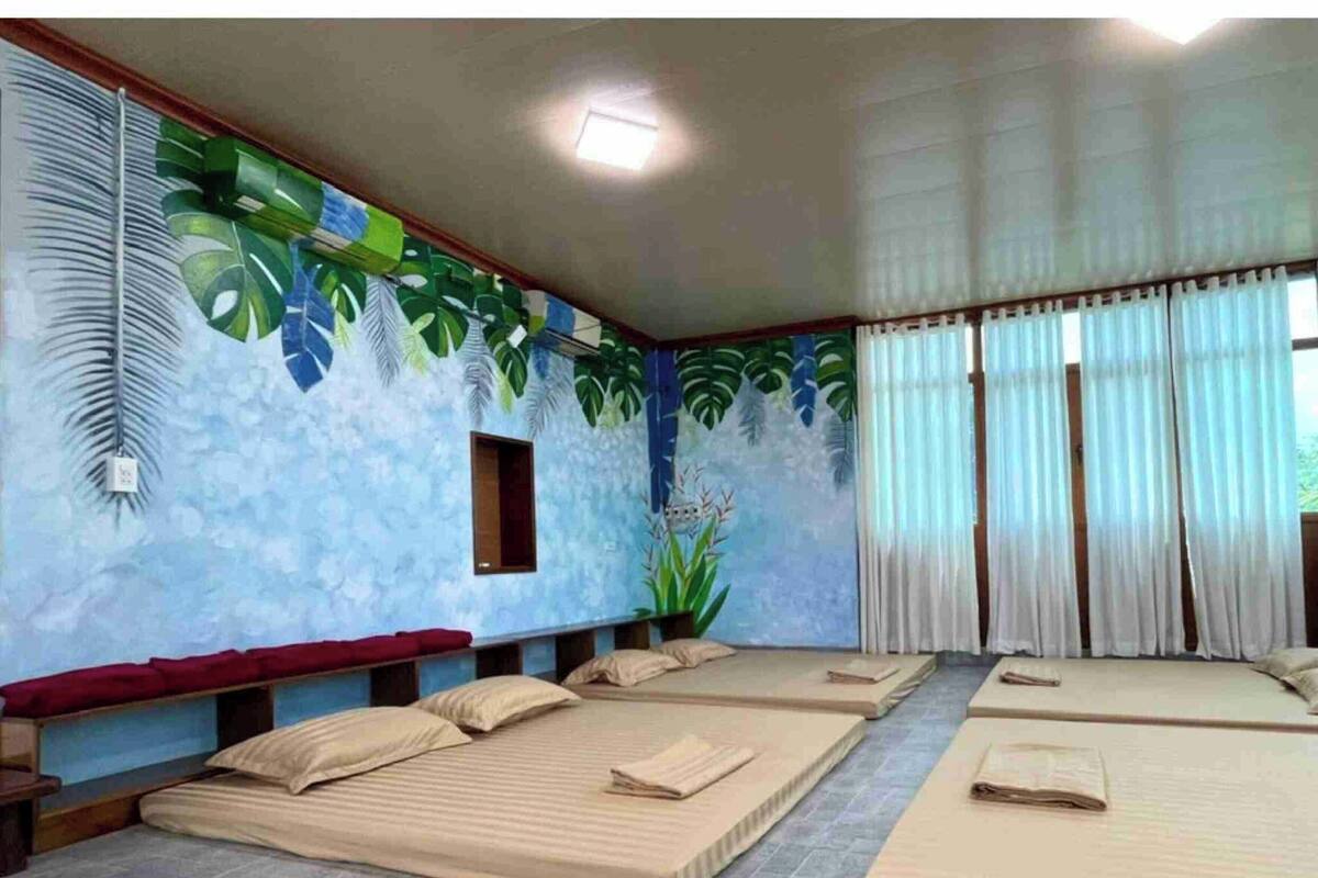 Quarto com quatro camas, cada uma com colchão e almofadas. Há uma pequena janela e uma porta na parede.