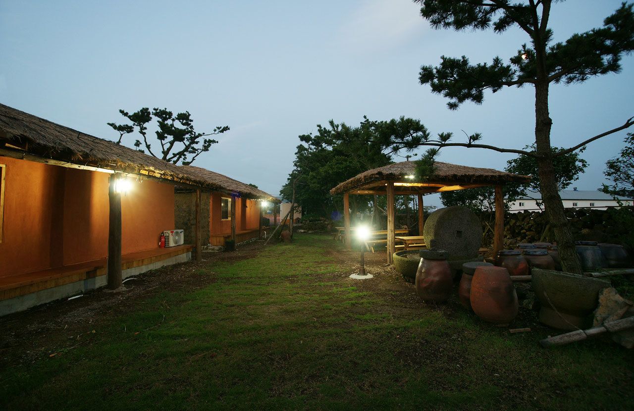 Ambiente rústico ao ar livre com construções de palha, área de estar coberta e panelas de cozinha tradicionais.