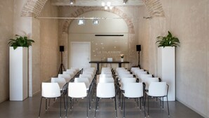 Sala de conferência com cadeiras brancas dispostas em fileiras, uma mesa preta com microfone e duas plantas em vasos.