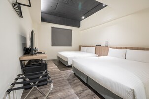 Quarto de hotel com duas camas, uma mesa de escritório com computador, tela de projetor e teto com vigas aparentes.