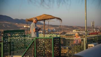 Um homem está debaixo de um guarda-sol de palha, uma mulher numa cabine verde, e uma vista de montanhas e edifícios ao longe.
