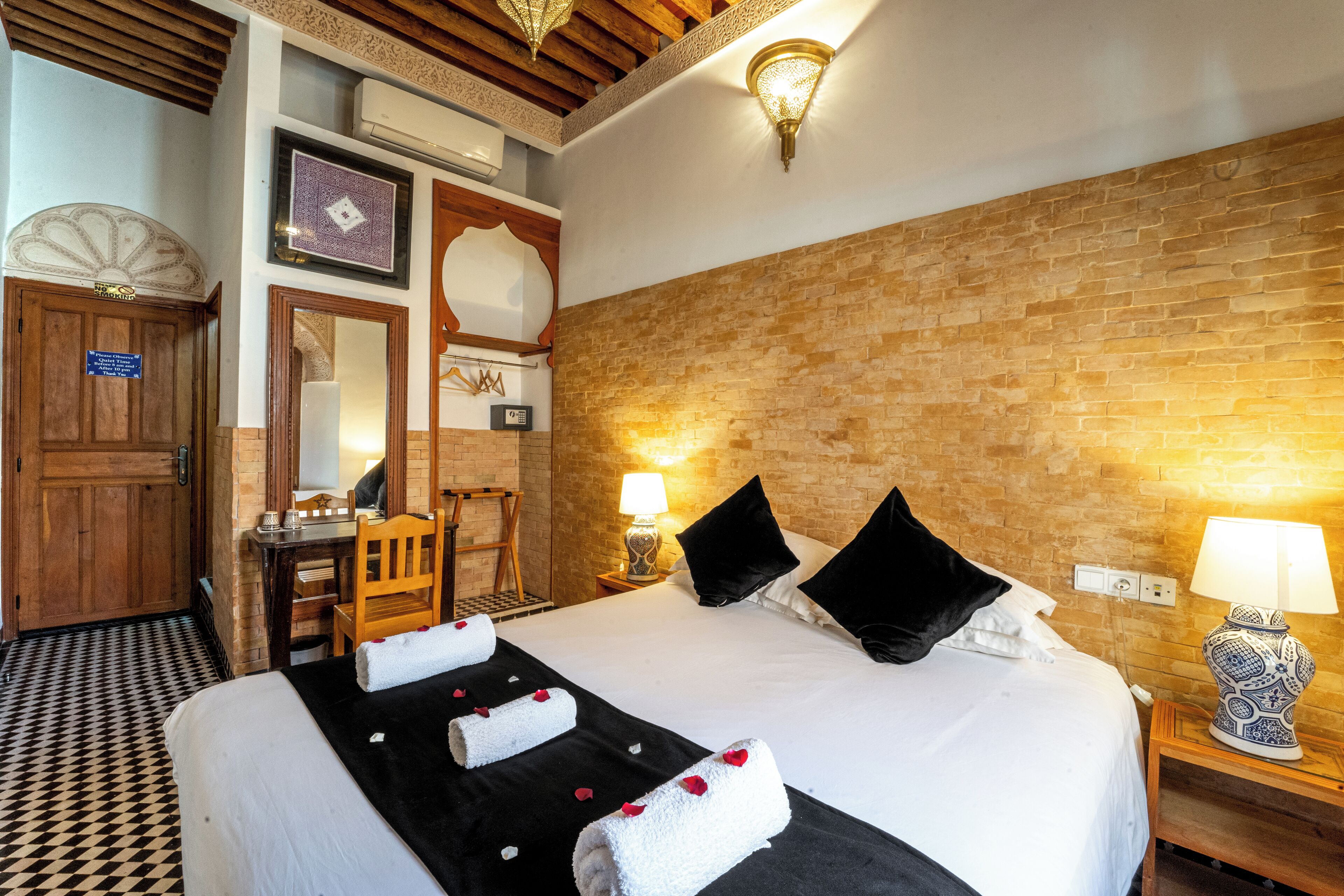 Quarto de hotel com uma cama grande, uma escrivaninha de madeira, uma cadeira, um espelho, parede de tijolos e um lustre.