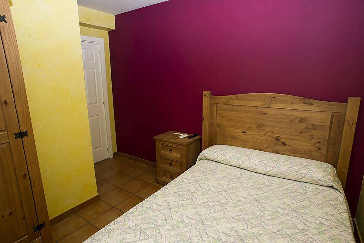 Quarto com cama de madeira, mesa de cabeceira, gaveteiro e porta que dá para outro cômodo.